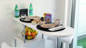 Disposição para o café da manhã com uma cesta de frutas, caixa de cereais e bebidas sobre uma mesa branca.