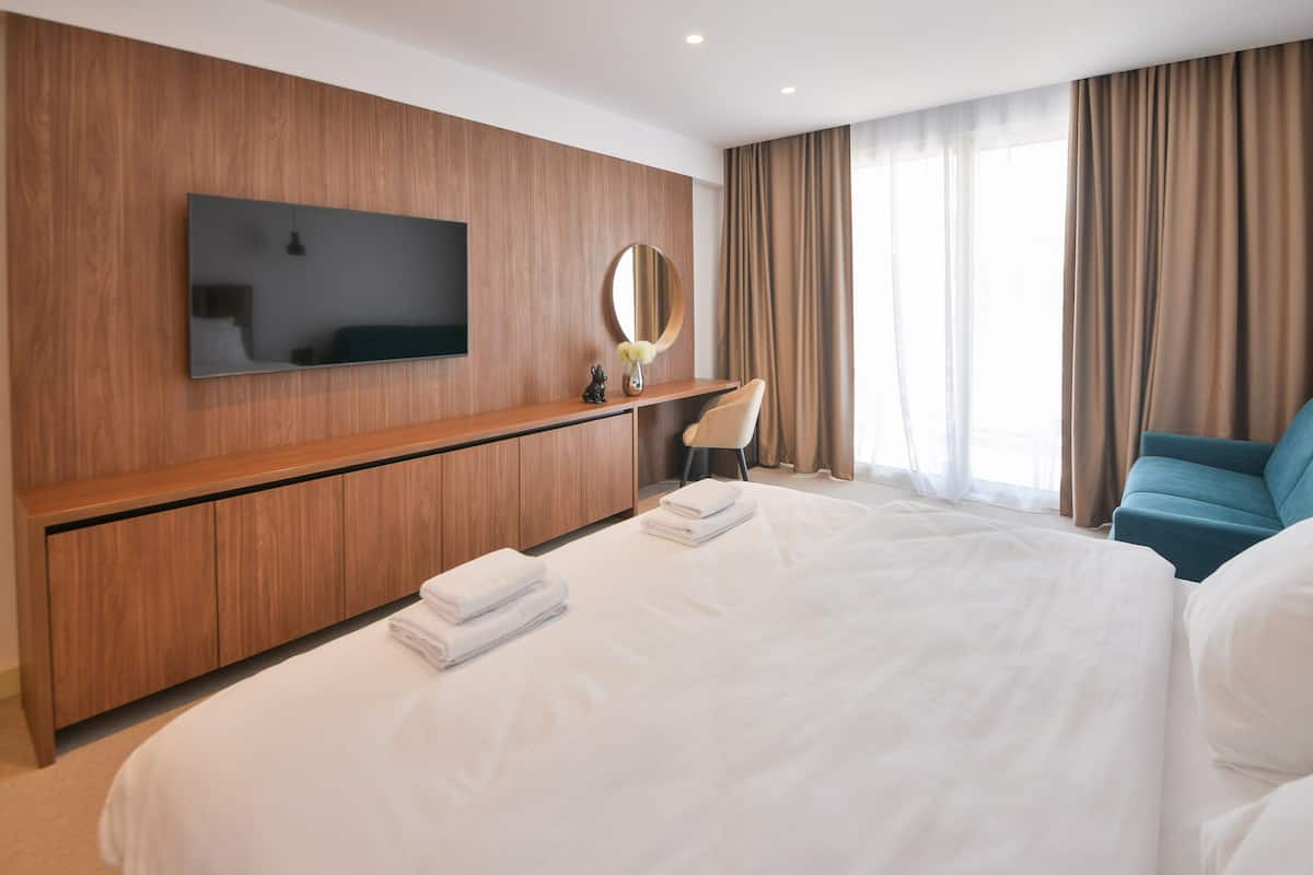 Quarto de hotel moderno com uma cama grande, uma TV de tela plana fixada numa parede revestida de madeira, uma pequena mesa com uma cadeira e vista para o exterior através de amplas janelas.