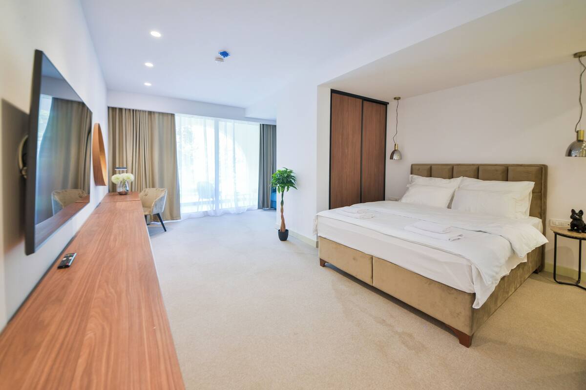 Quarto de hotel moderno com uma cama grande, uma escrivaninha de madeira, televisão de tela plana e uma grande janela com cortinas.