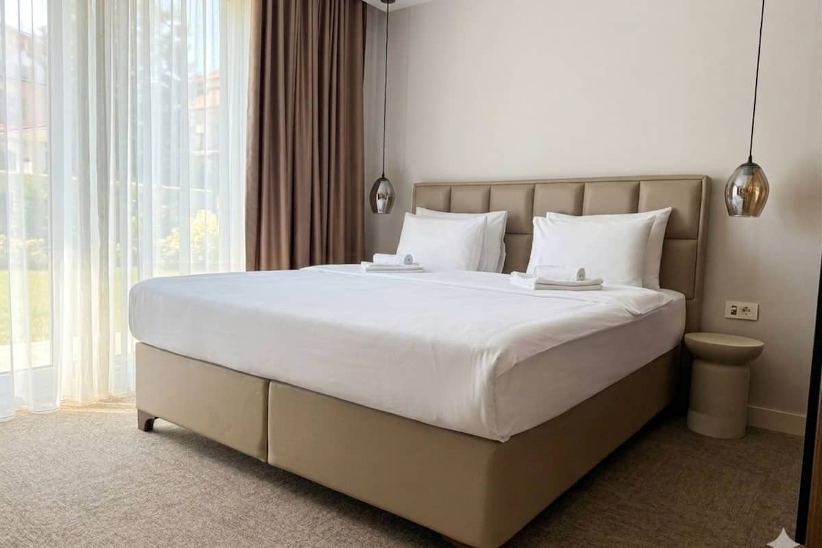Quarto de hotel com uma cama grande, uma mesa de cabeceira, uma luminária e vista para o exterior.