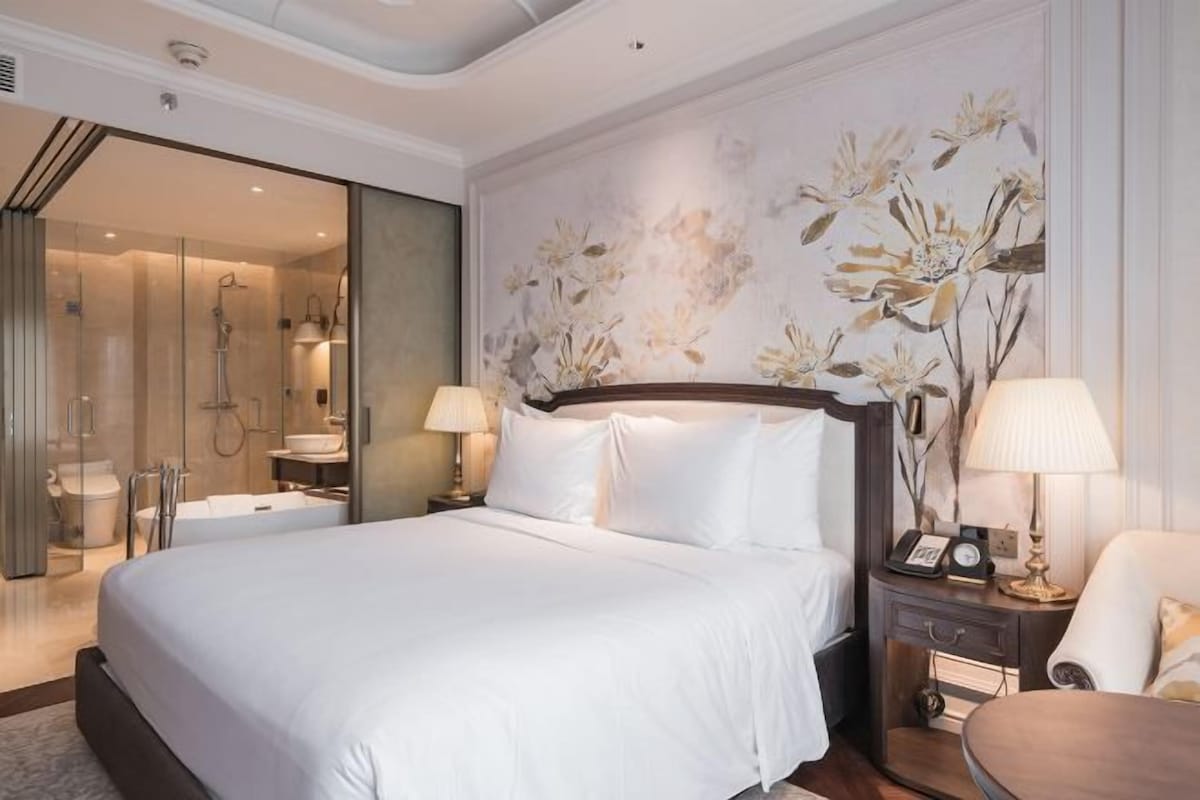 Quarto de hotel com uma cama grande, uma mesa de cabeceira com abajur, uma cadeira, área de chuveiro e parede com um mural floral.