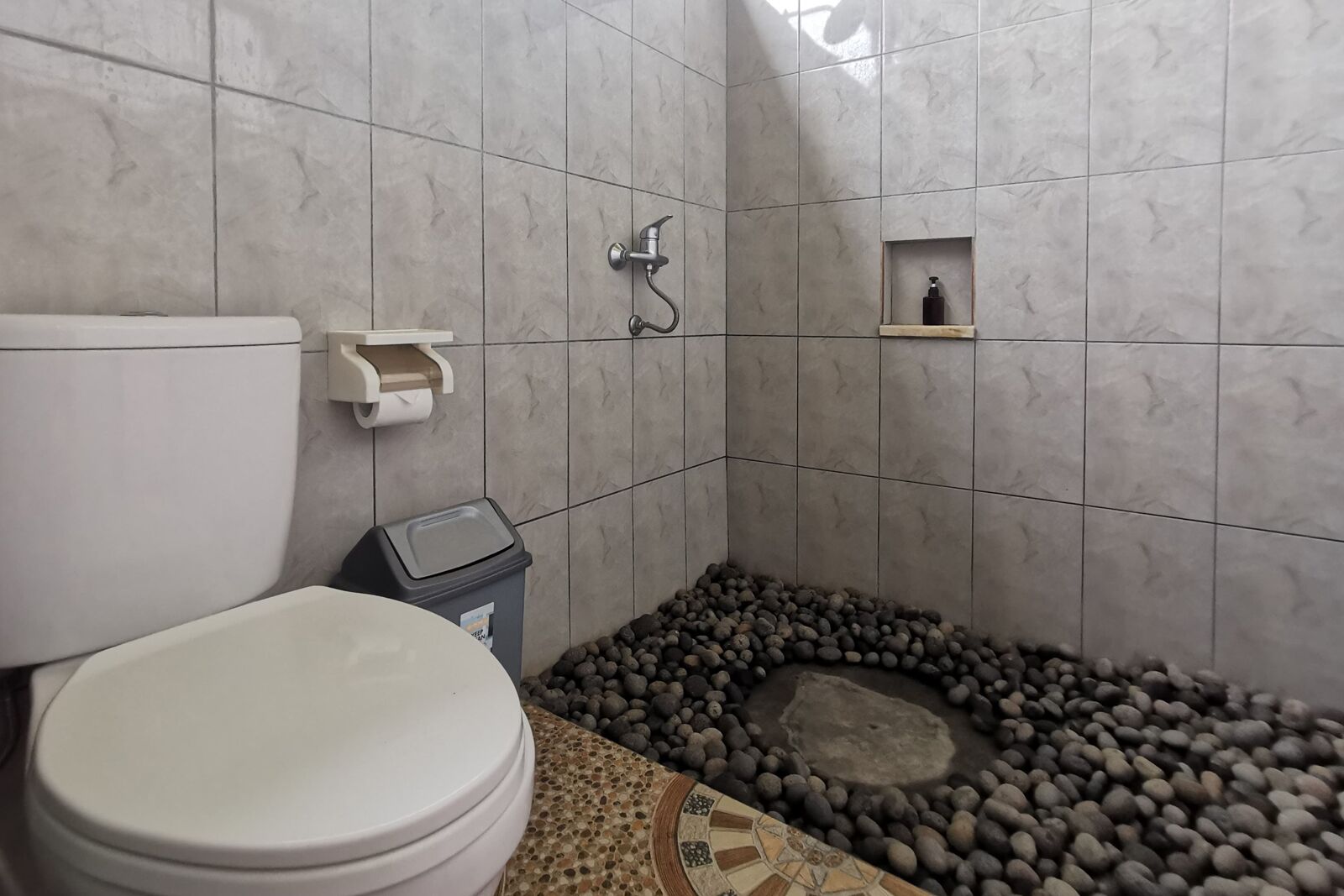 Casa de banho com sanita, um contentor de lixo, zona de duche com chão de pedras e uma pequena estante com uma garrafa.