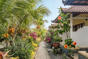 Caminho ladeado por vegetação exuberante e plantas floridas, conduzindo a um edifício com telhado de telhas.