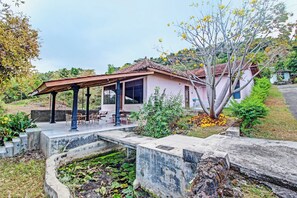 Uma casa com alpendre, um jardim com pequeno lago e uma árvore com flores amarelas.