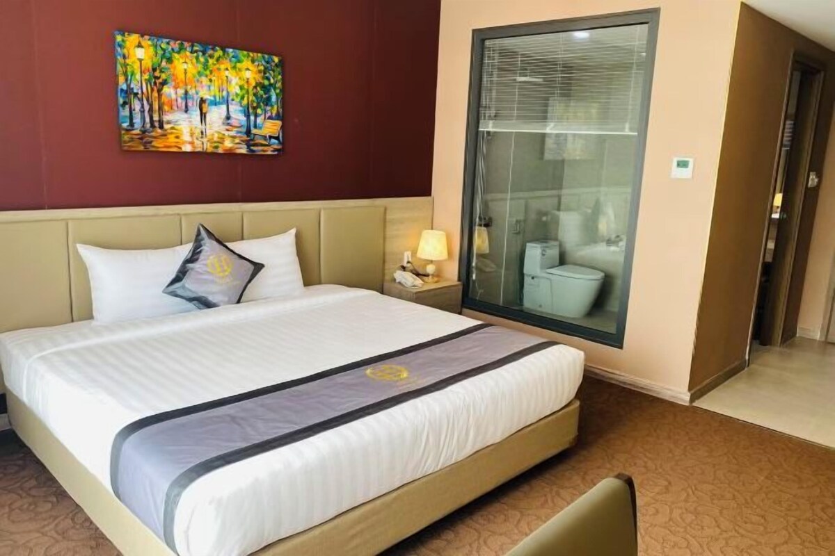 Quarto de hotel com cama, mesa de cabeceira, um quadro na parede e banheiro visível por uma porta de vidro.