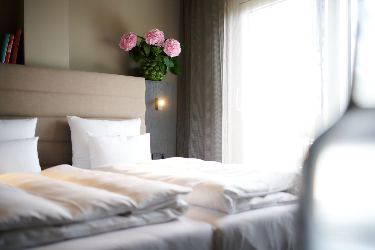Quarto de hotel com cama, almofadas, luminária na parede e um vaso com flores.
