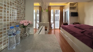 Quarto de hotel com cama, televisão, janela com cortinas, uma mesinha com um vaso de flores e duas garrafas de água.