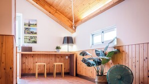 Interior de madeira com cozinha, dois banquinhos, um ventilador e um vaso de planta.