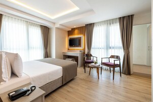 Quarto de hotel com uma cama, uma escrivaninha com televisão, uma cadeira e uma mesinha de centro com um vaso.