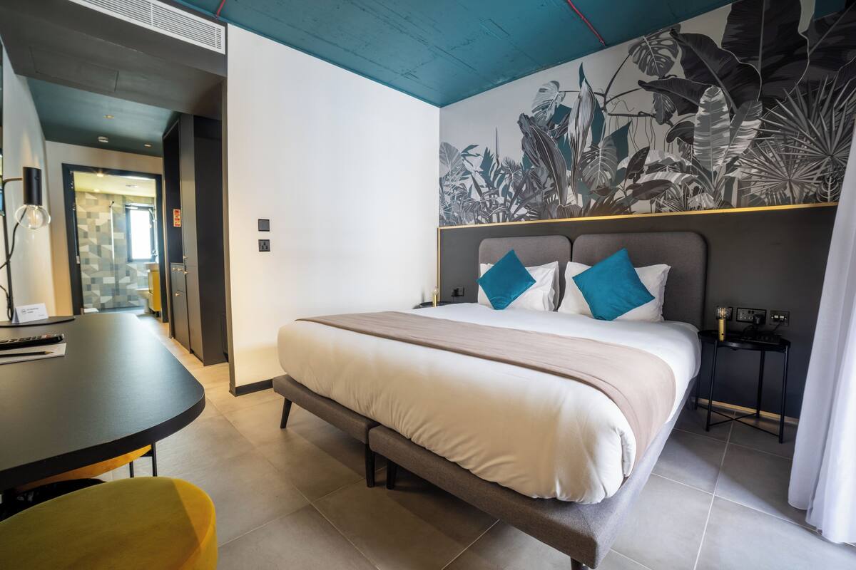 Quarto de hotel moderno com uma cama grande, uma pequena mesa, um banco e um mural na parede.