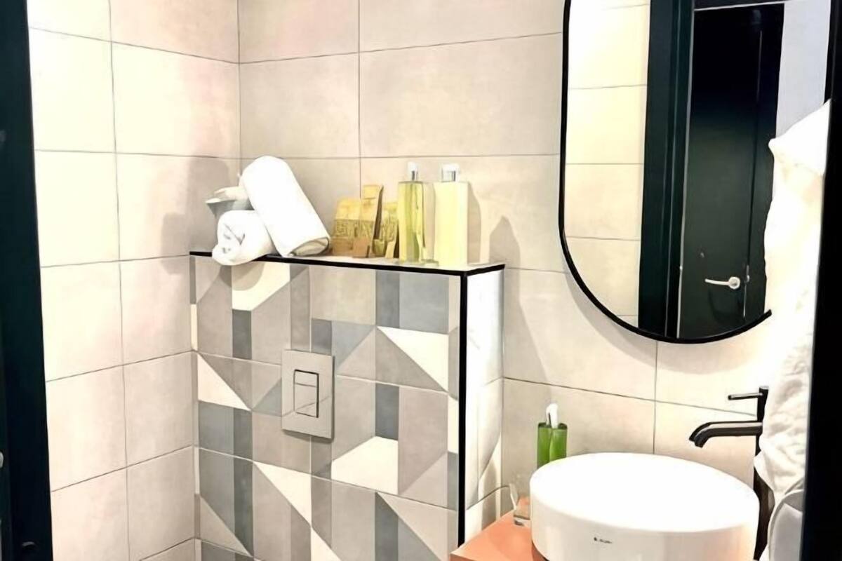 Um banheiro com design moderno, com um padrão geométrico de azulejos nas paredes, uma prateleira de vidro com produtos de higiene pessoal, uma pia branca e um espelho redondo.