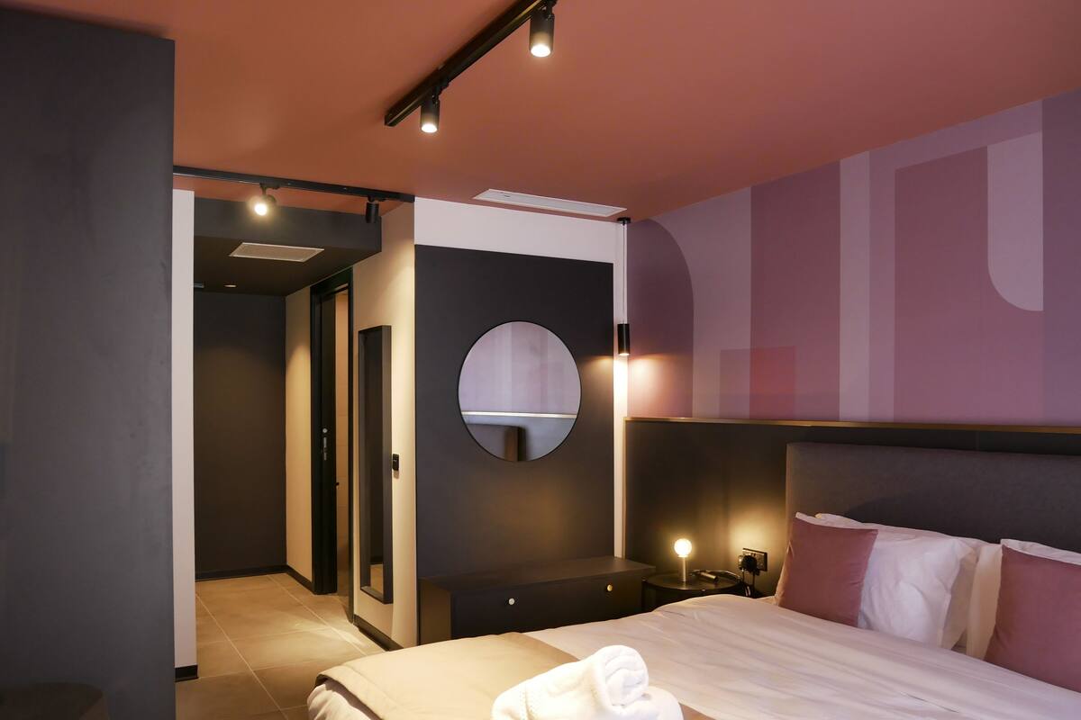 Quarto de hotel moderno com uma cama grande, um espelho redondo, mesa de cabeceira com abajur e vista para o corredor.