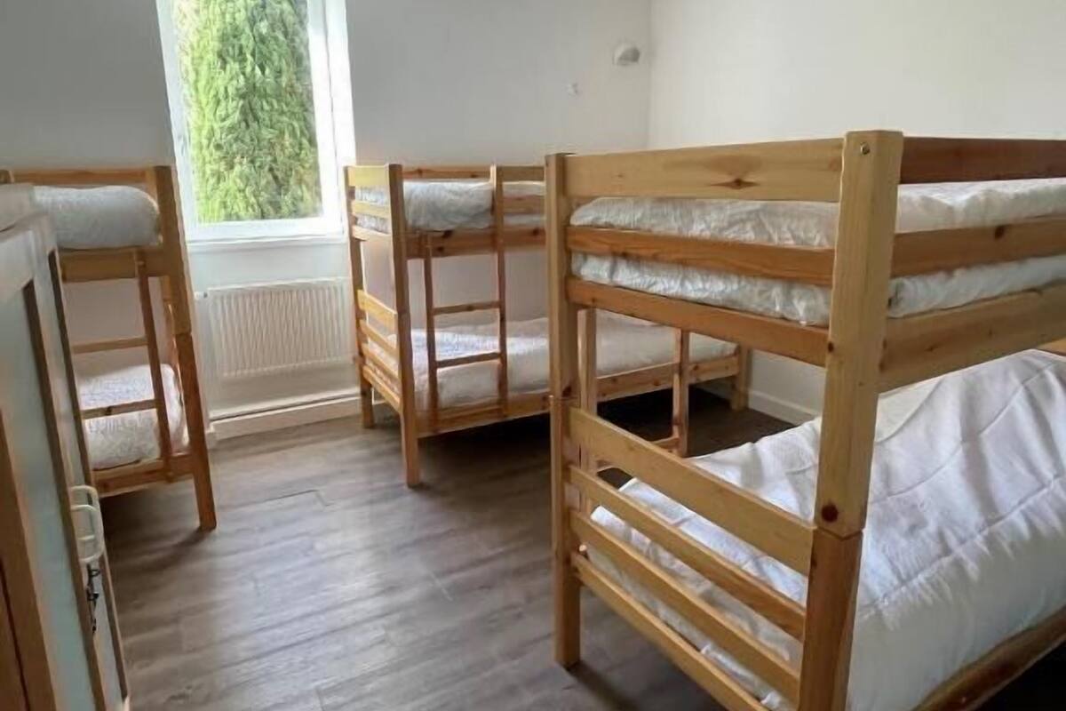 Quarto com três beliches, piso de madeira, janela com vista para vegetação e luminária no teto.