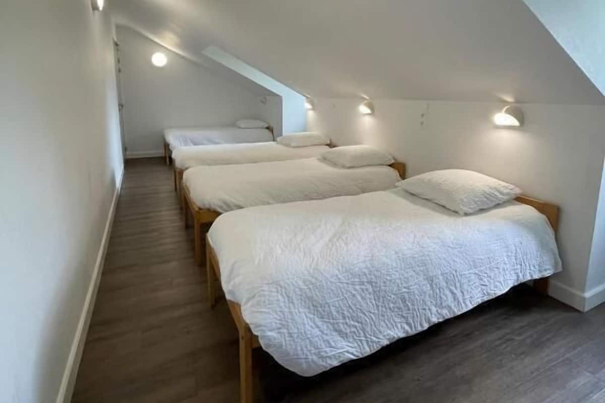 Quarto com teto baixo e inclinado, quatro camas alinhadas na parede, piso de madeira e iluminação embutida no teto.