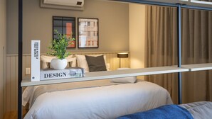 Quarto de hotel moderno com uma cama, uma mesa de cabeceira com uma planta, um quadro na parede e uma janela grande com cortinas.