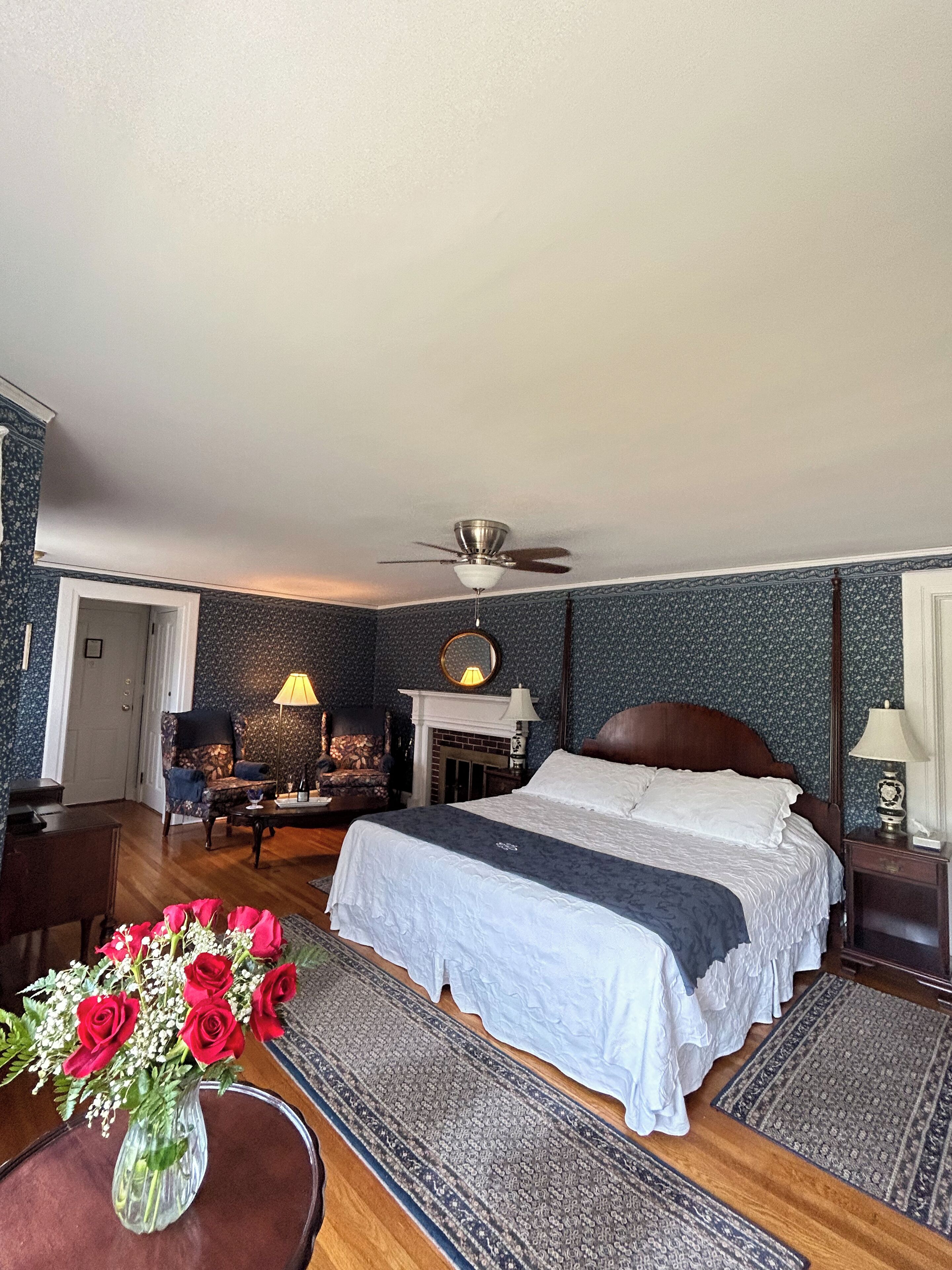 Quarto de hotel com uma cama grande, um ventilador de teto, lareira, escrivaninha e uma cadeira.