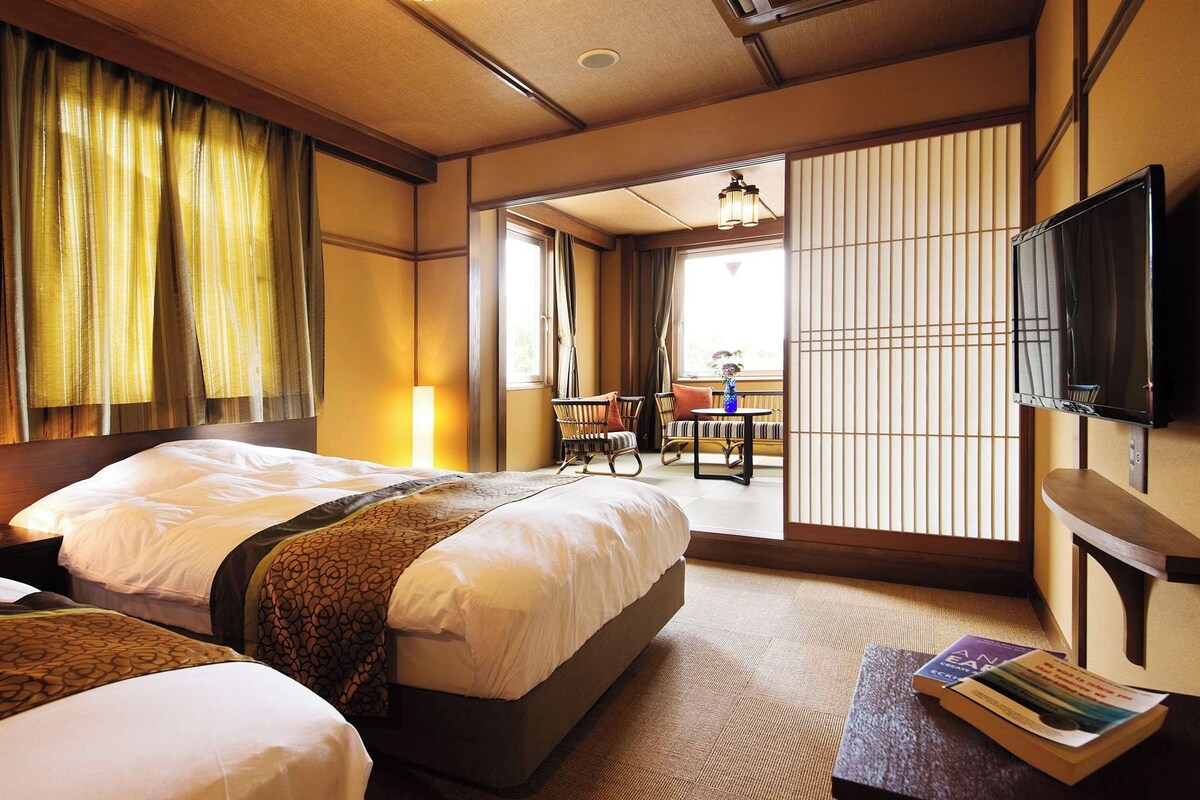 Quarto de hotel com duas camas, uma televisão, uma estante e vista para o exterior.