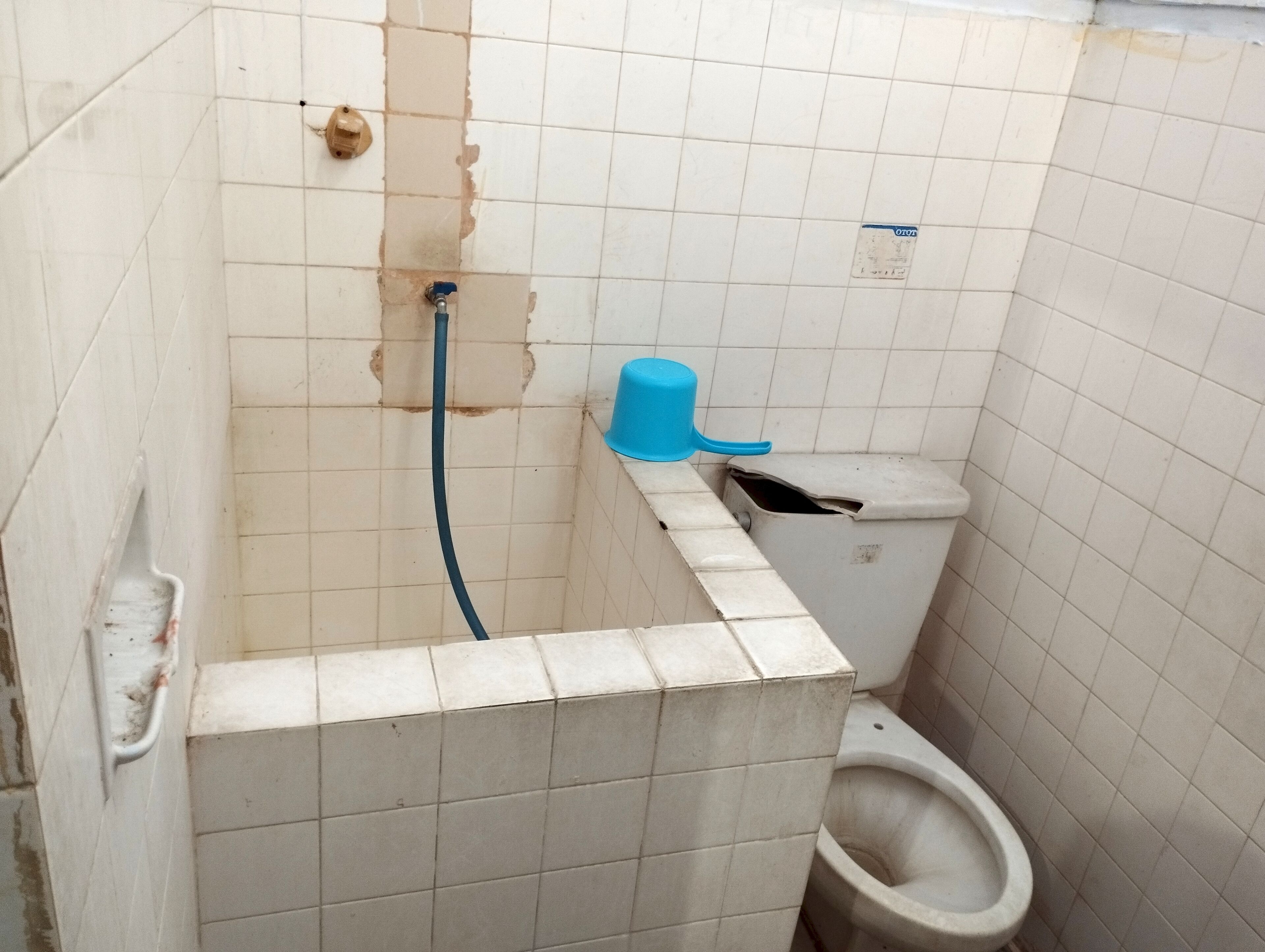 Um banheiro pequeno, com azulejos, um vaso sanitário, um balde azul e uma mangueira de chuveiro.