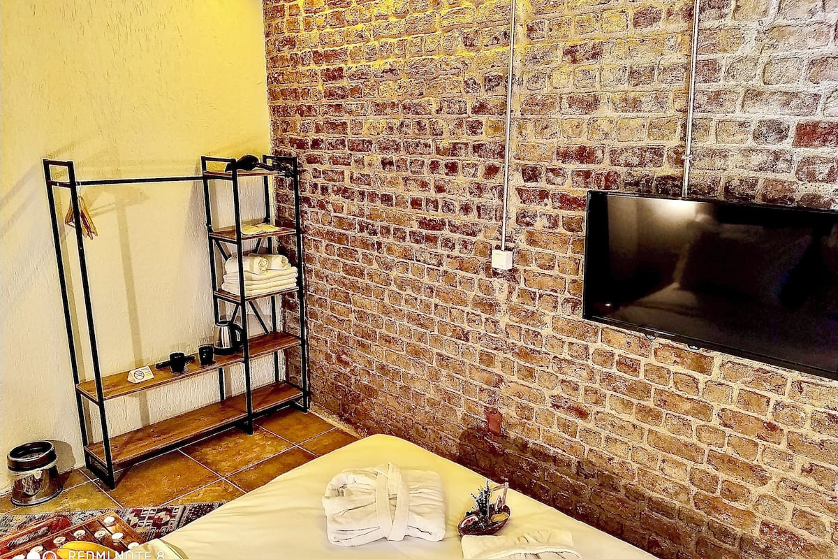 Quarto com parede de tijolos, televisão fixada nela, prateleira com toalhas e cama com lençol branco.