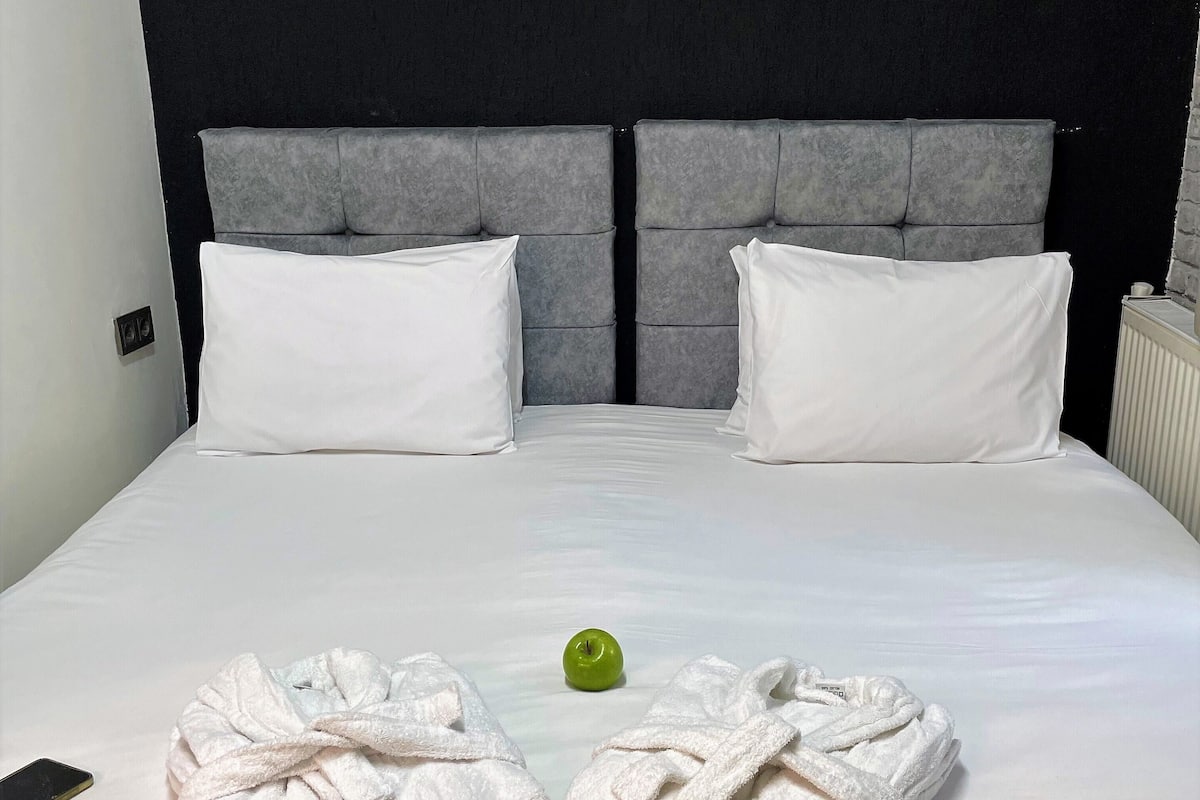 Cama bem arrumada com duas almofadas, uma maçã verde e duas toalhas sobre um cobreleiro branco.