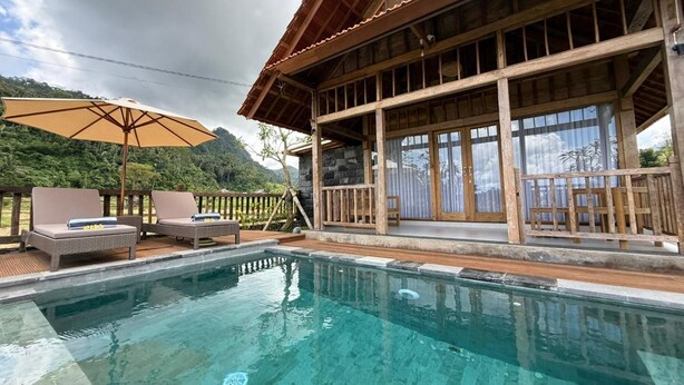 Uma área à beira da piscina com espreguiçadeiras e um guarda-sol, um deck de madeira e um edifício de pedra com grandes janelas.