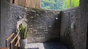 Área de chuveiro externo com paredes de pedra, um banco de madeira, uma planta em vaso e vista para o verde.