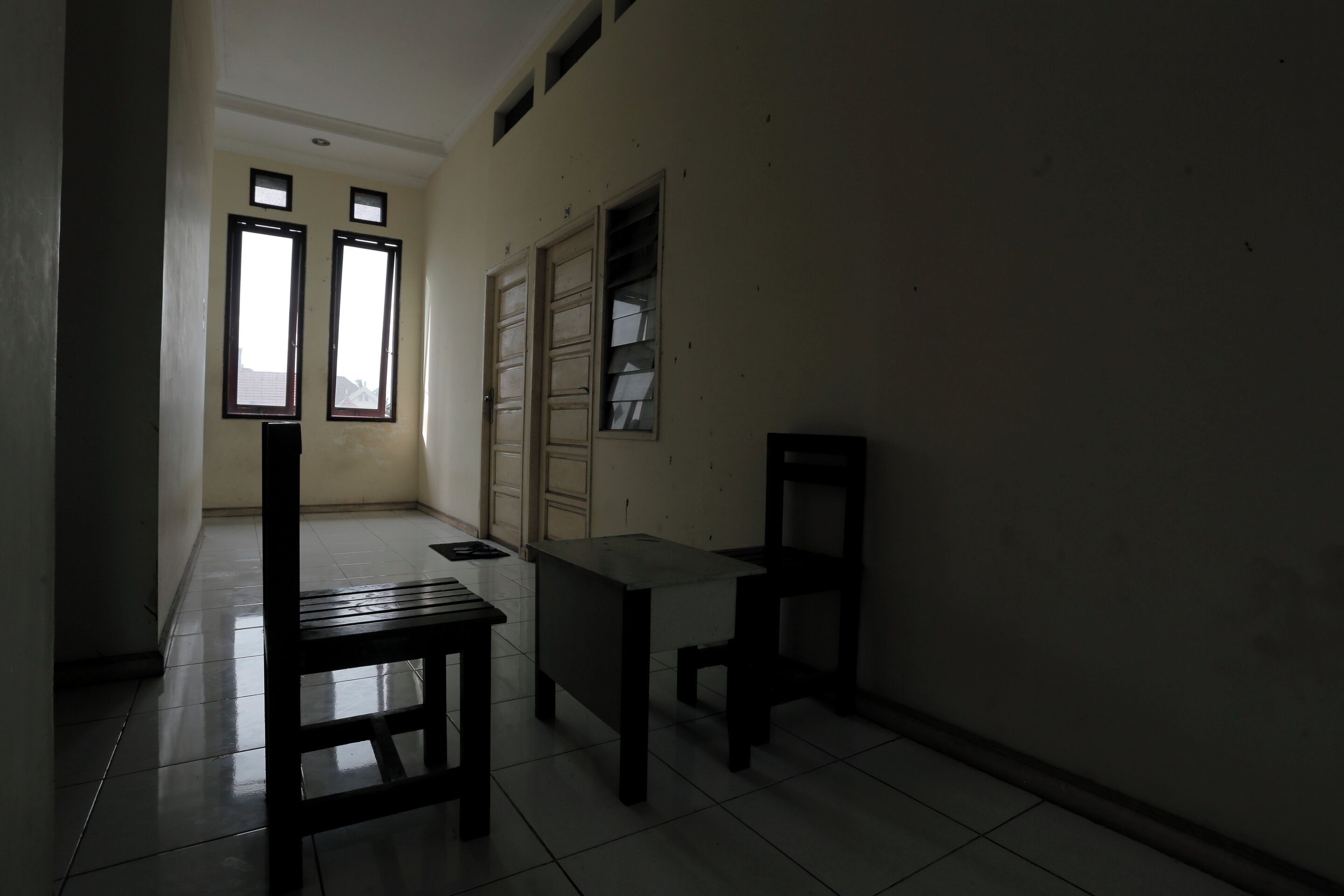 Um corredor com uma cadeira de madeira, uma pequena mesa e um guarda-roupa. Há duas janelas com persianas e uma porta com espelho.