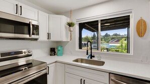 Cozinha moderna com armários brancos, eletrodomésticos de aço inoxidável, janela com vista e planta pendurada.