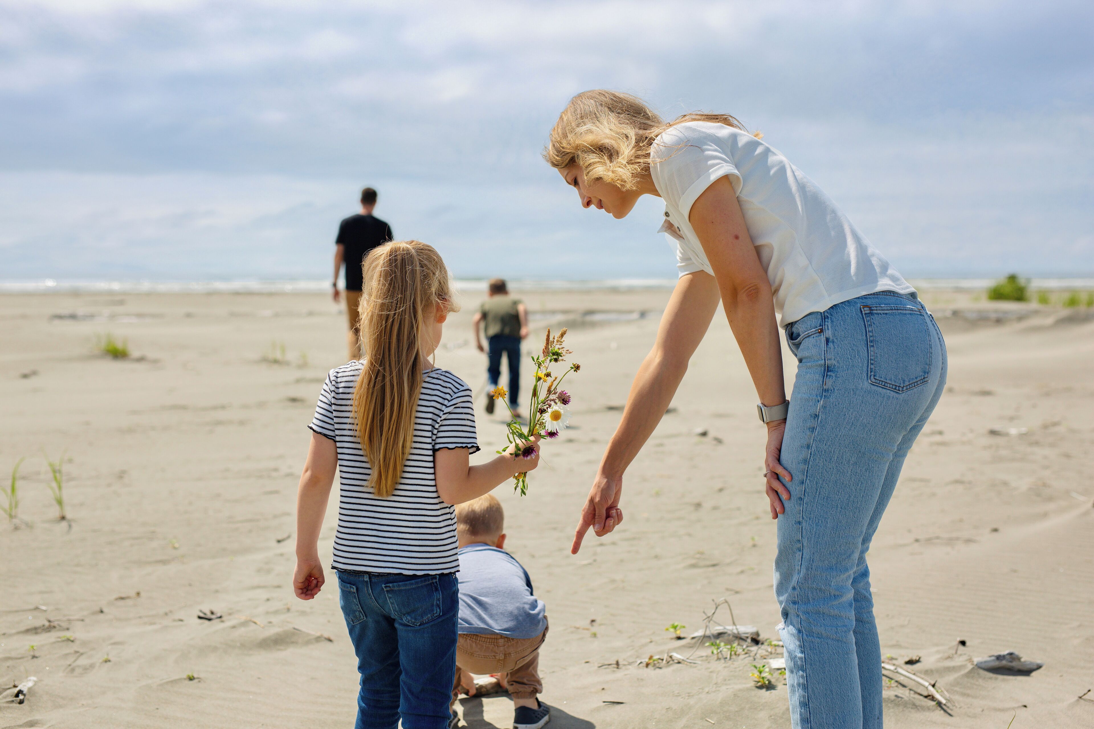 Uma praia de areia com pessoas, incluindo uma mulher curvada sobre uma criança segurando flores.