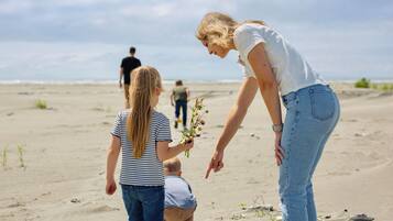 Uma praia de areia com pessoas, incluindo uma mulher curvada sobre uma criança segurando flores.