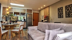 Uma sala moderna com um sofá cinzento, mesa de jantar de madeira e cadeiras. Uma cozinha com micro-ondas e frigorífico é visível ao fundo.