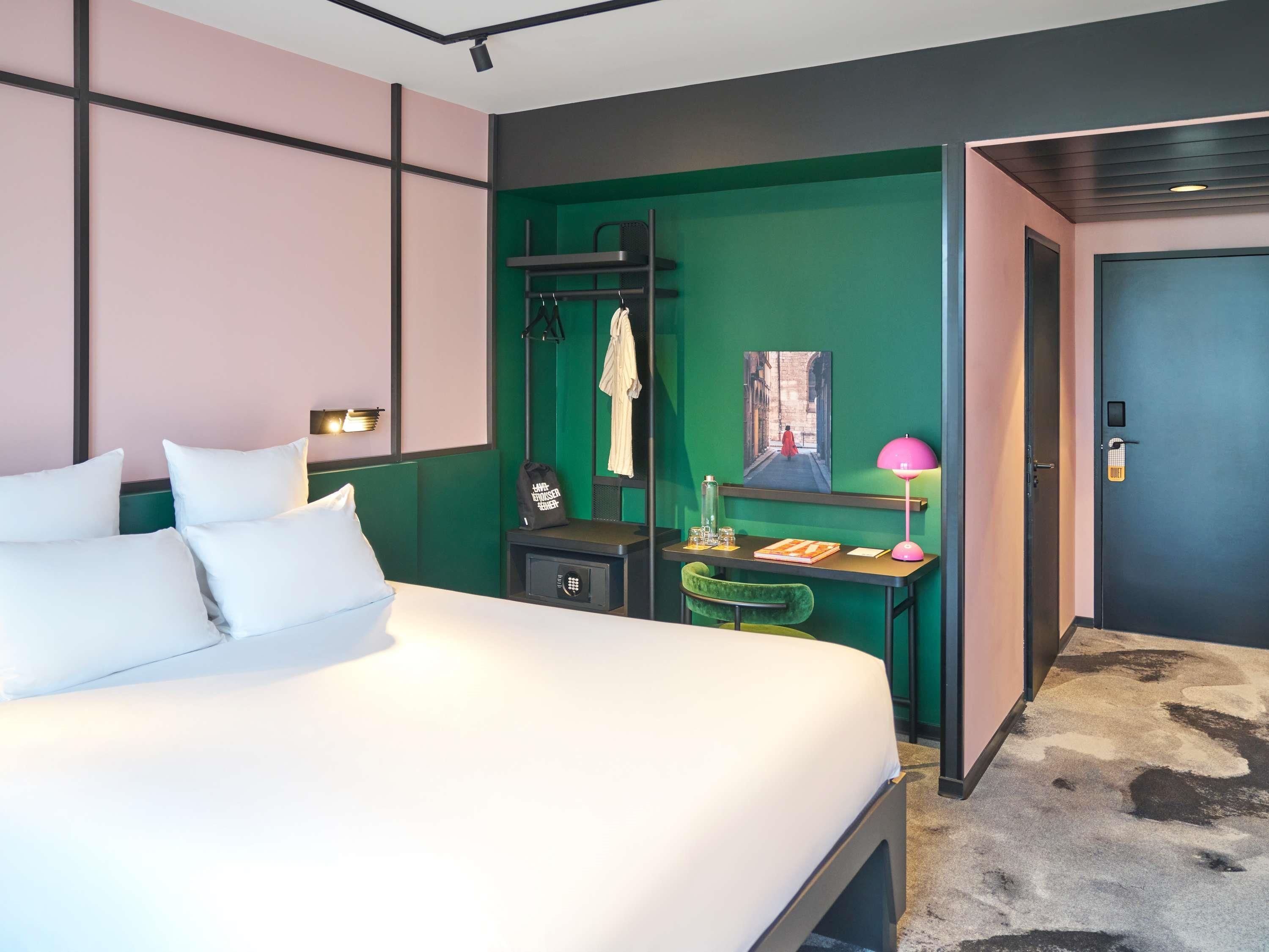 Quarto de hotel moderno com uma cama grande, uma escrivaninha com abajur, uma parede verde decorativa e uma porta com fechadura.