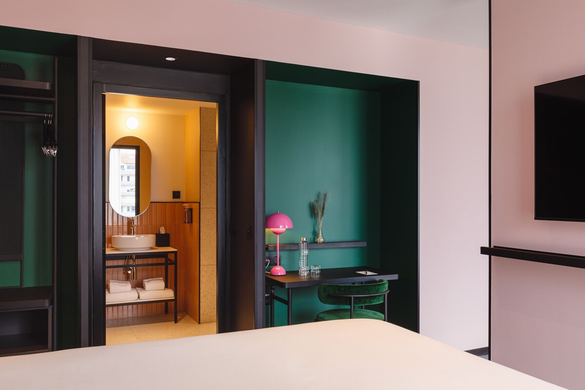 Quarto de hotel moderno com cama, escrivaninha com cadeira, mesinha de centro com abajur e espelho.