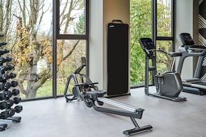 Uma academia com equipamentos de cardio, halteres e uma grande janela com vista para árvores.