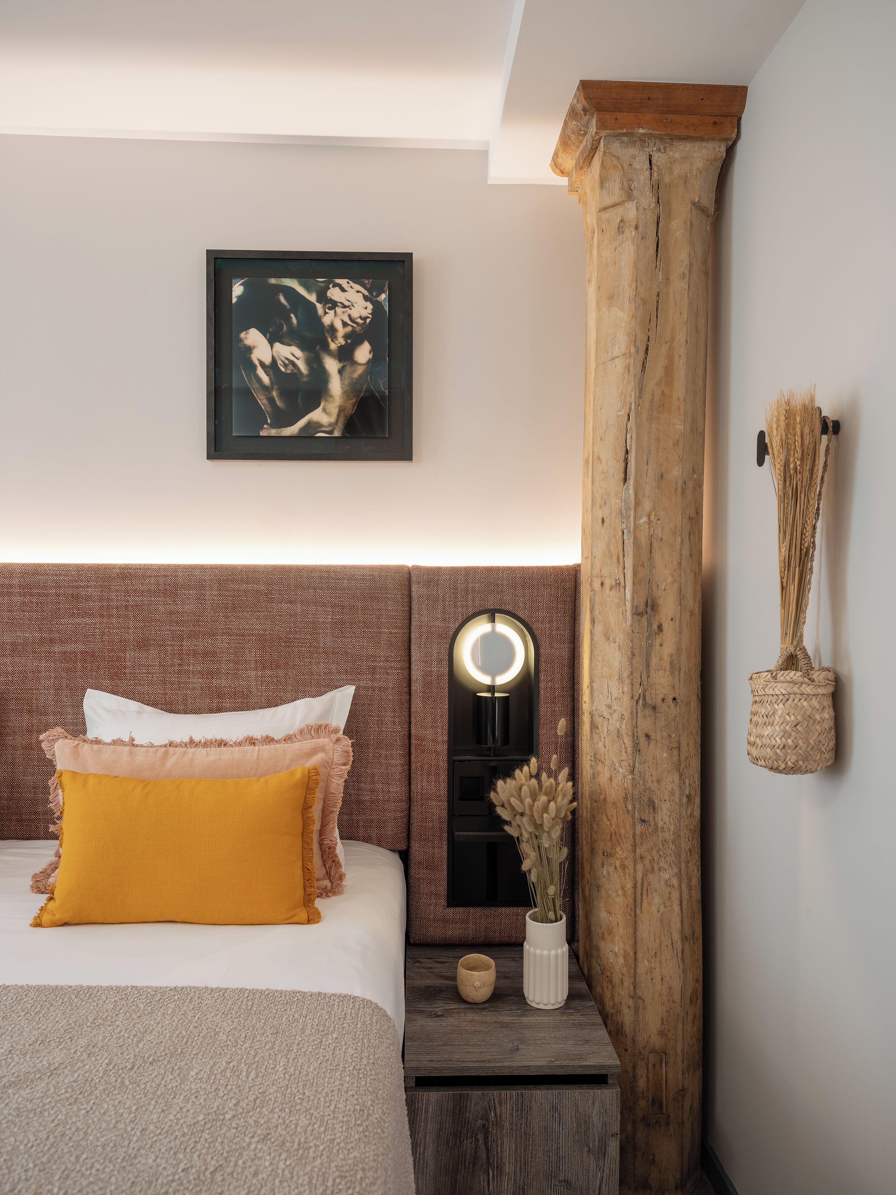 Quarto com cama, mesa de cabeceira com abajur, quadro na parede e um pilar de madeira.