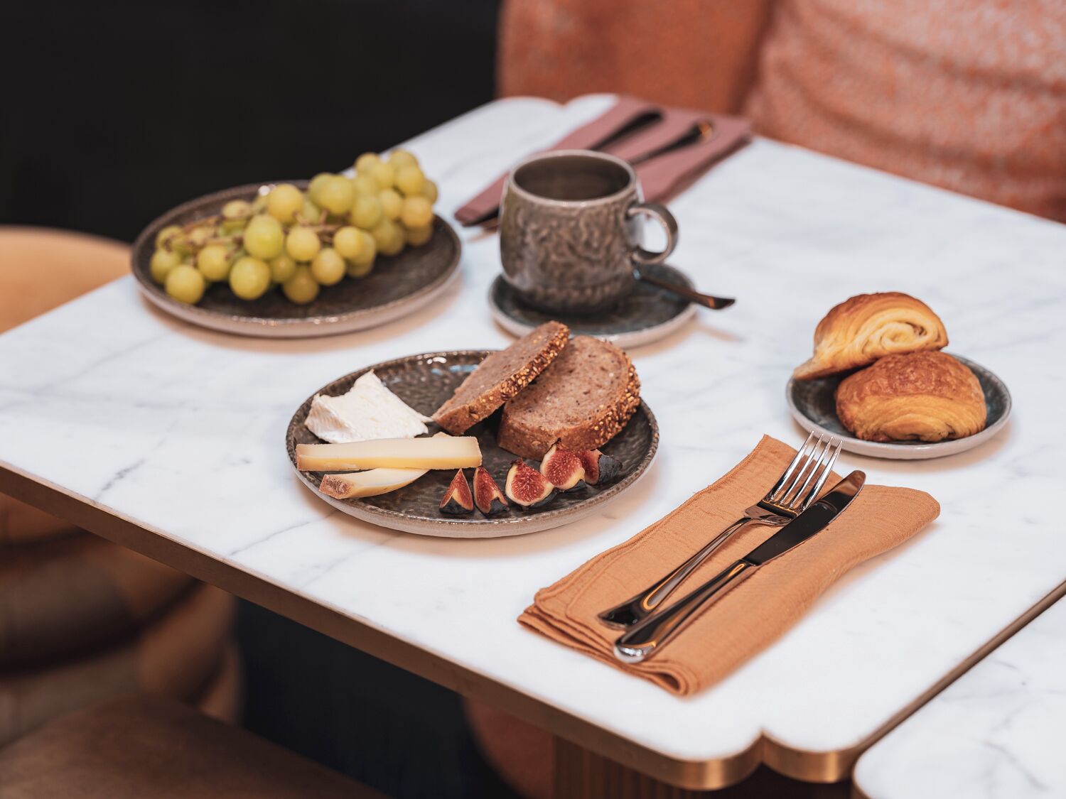 Uma mesa de mármore com pratos de queijo, pão, figos, pastelaria e uma chávena de café, além de talheres.