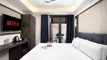 Quarto de hotel moderno com uma cama grande, televisão de tela plana na parede, mesa de cabeceira com chaleira, xícaras e um livro, e vista para uma varanda com cortinas.