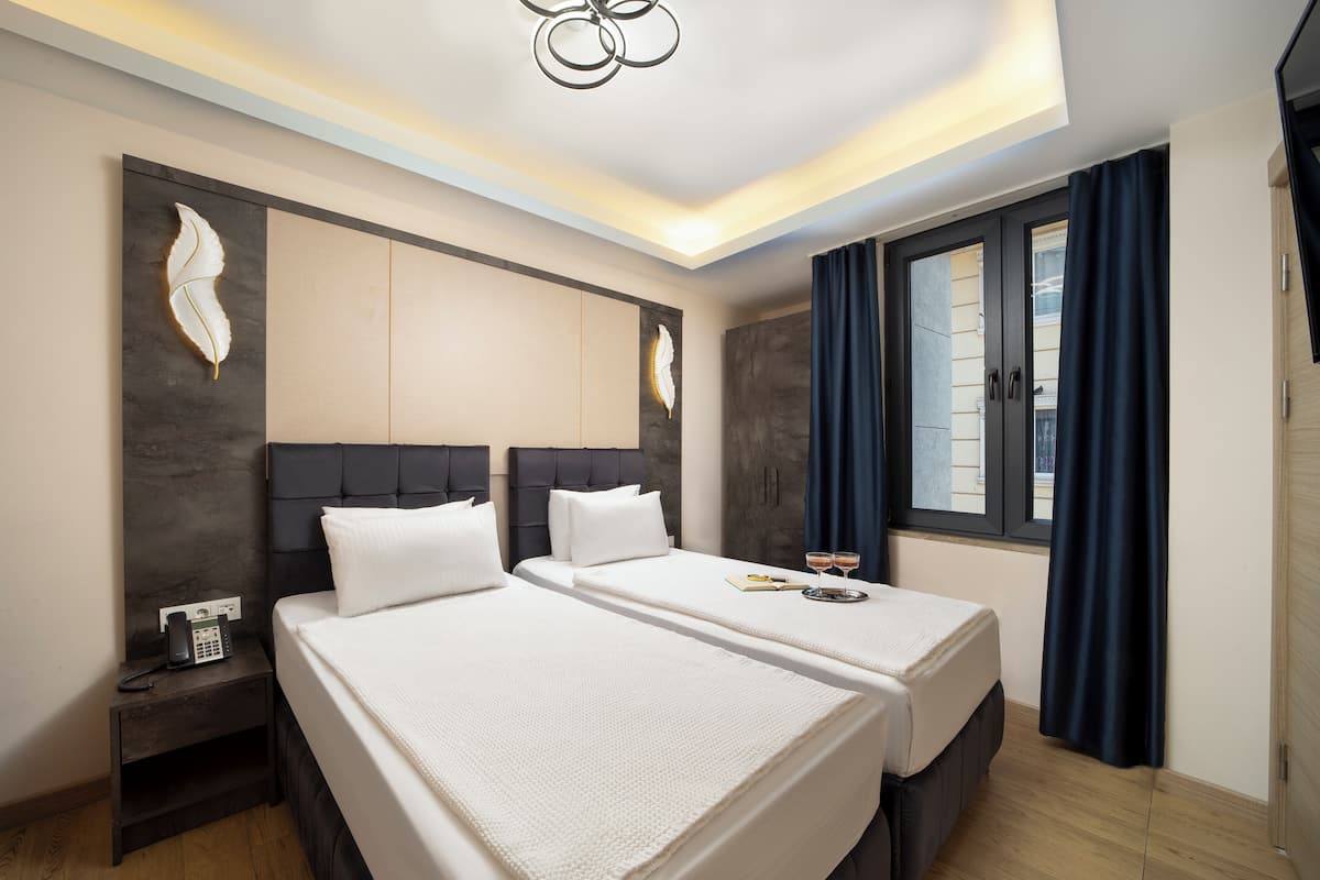 Quarto de hotel com duas camas, cabeceira escura, telefone fixado na parede e janela com cortinas.