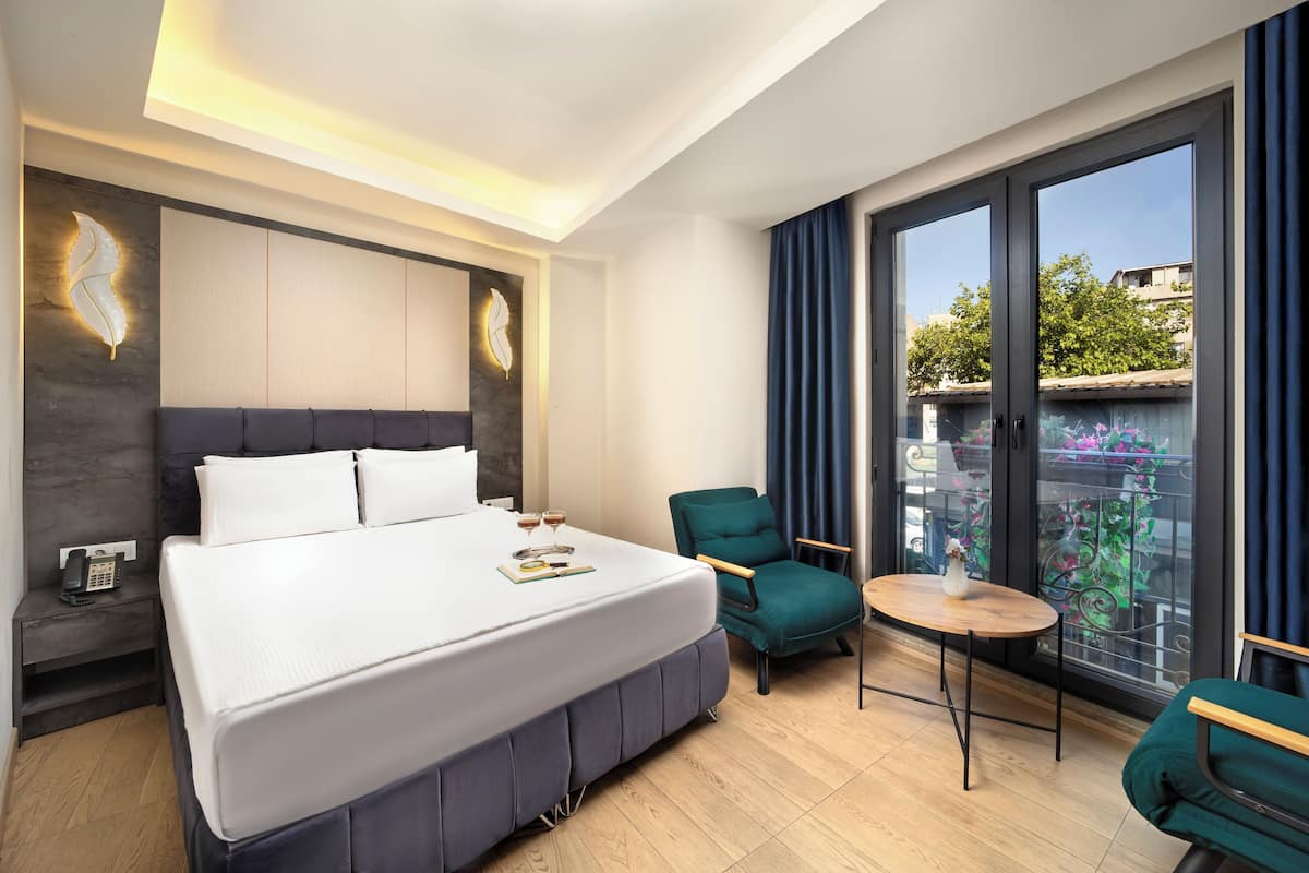 Quarto de hotel moderno com uma cama grande, uma mesa redonda pequena, uma poltrona verde e uma varanda com vista para a cidade.