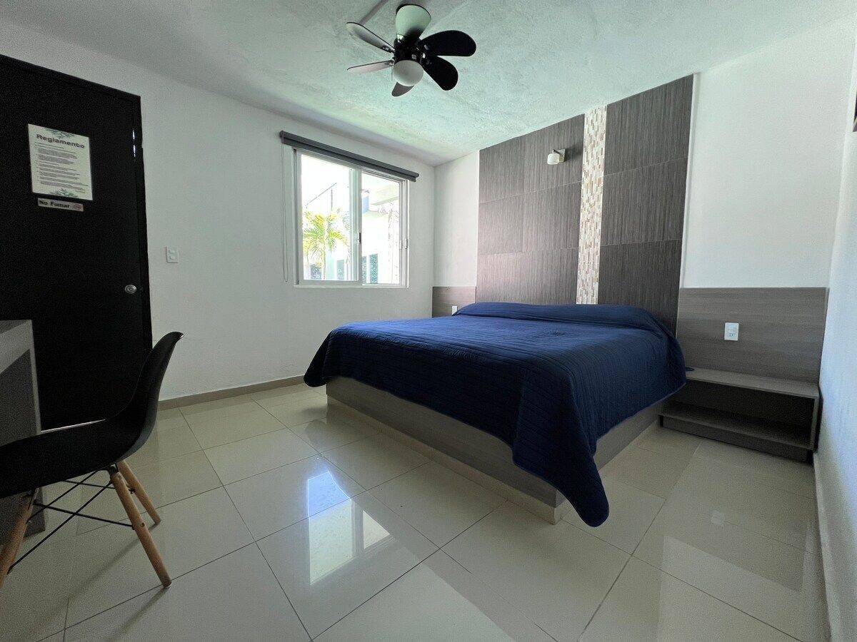 Quarto com cama, escrivaninha, cadeira, janela e ventoinha no teto.
