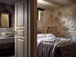 Quarto com parede de pedra, cama com colcha branca, porta de madeira e banheiro com pia de mármore e espelho.