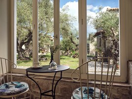 Quarto com vista para um jardim através de grandes janelas, uma mesa pequena com um vaso e duas cadeiras.