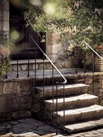 Escadaria de pedra com corrimão de metal que leva a uma porta, cercada por vegetação.