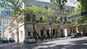Vista da rua com um edifício com várias sacadas, uma faixa de pedestres e carros estacionados.
