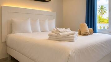Quarto de hotel com uma cama grande, roupa de cama branca, decoração com prancha de surf, ar condicionado e vista para palmeiras pela janela.