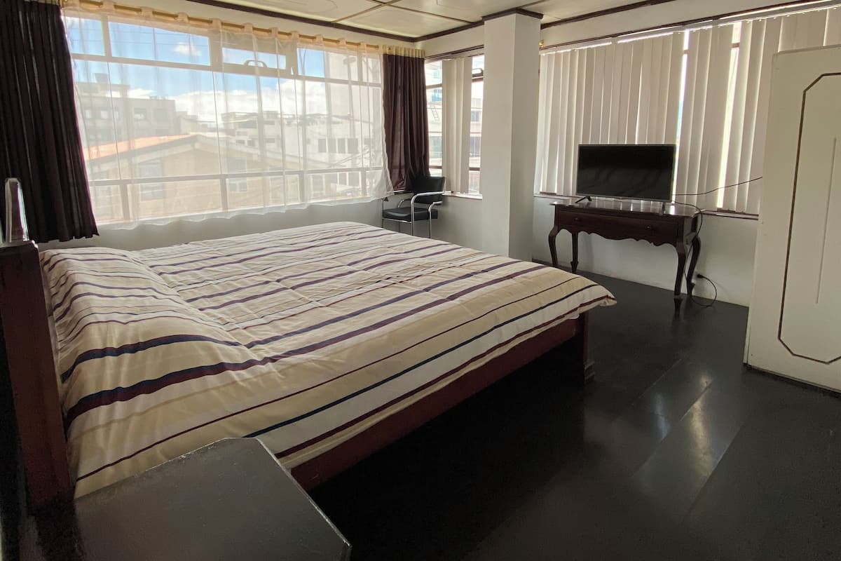 Quarto com cama grande, escrivaninha, televisão e vista para prédios pelas janelas.