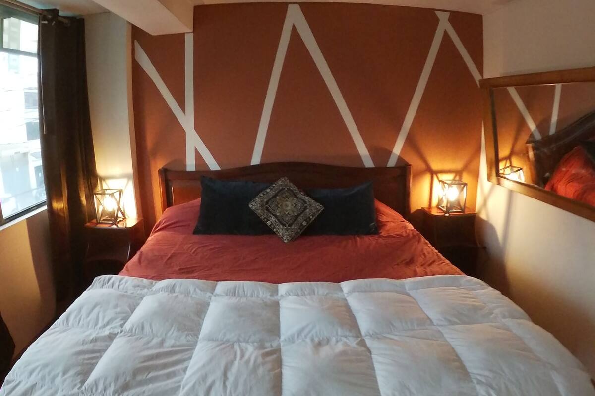 Uma cama com colcha branca, manta vermelha e um travesseiro decorativo. Há duas mesas de cabeceira com abajures. O quarto apresenta um design geométrico na parede e uma janela com cortinas.