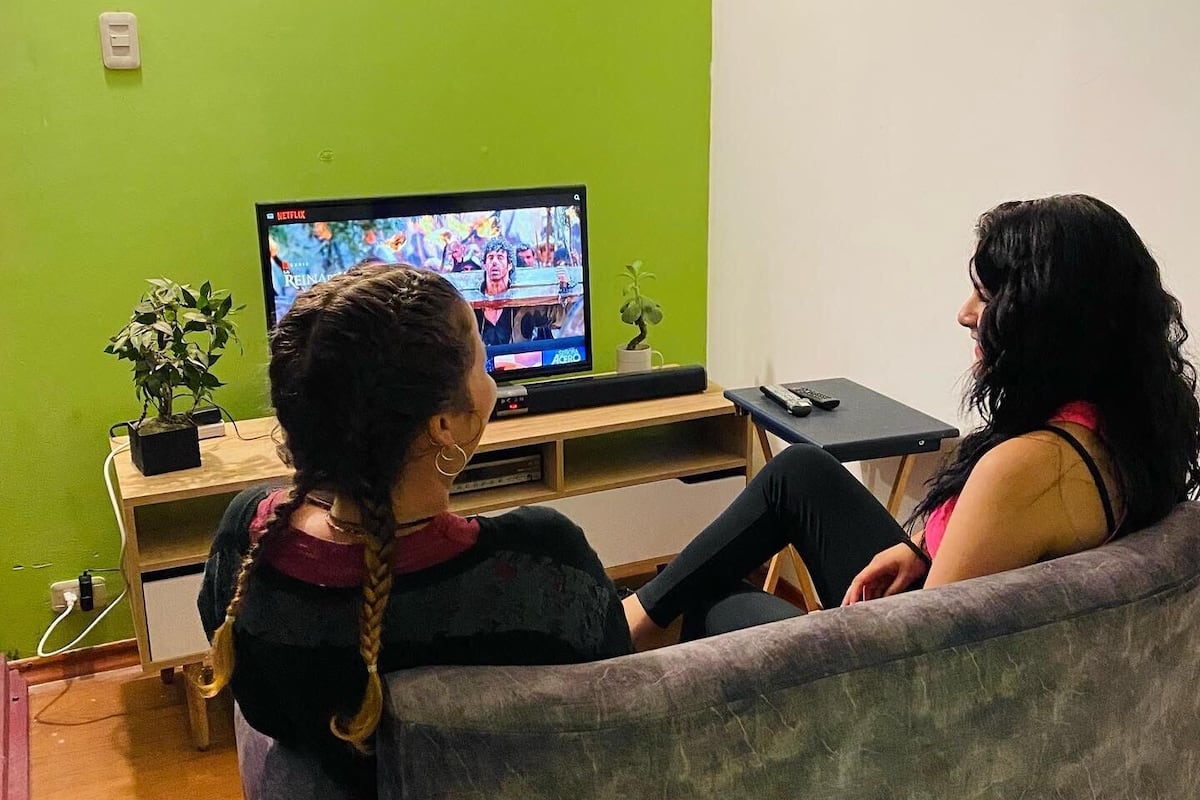 Uma sala de estar com parede verde, uma TV de tela plana e duas pessoas sentadas no sofá assistindo televisão.
