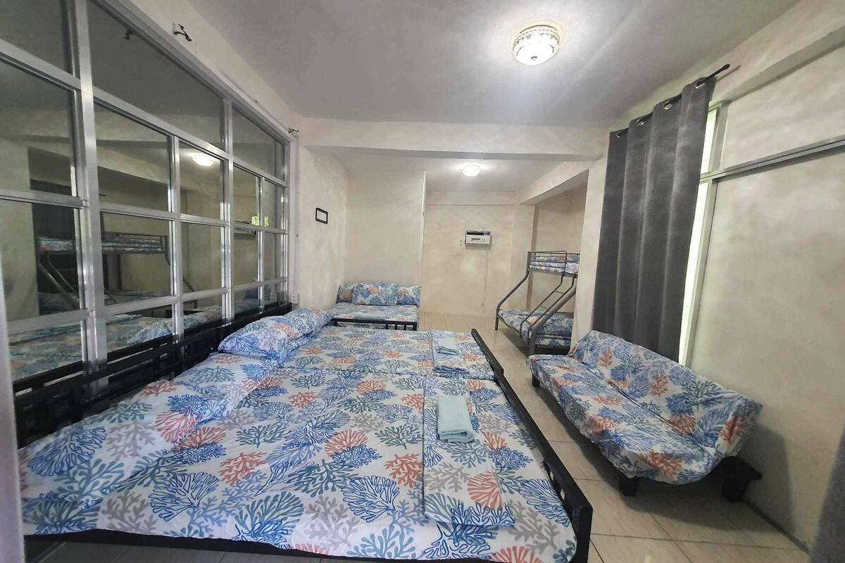 Quarto com uma cama grande, uma cama menor e um sofá. Há uma janela com cortinas, uma luminária no teto e um ar-condicionado fixado na parede.