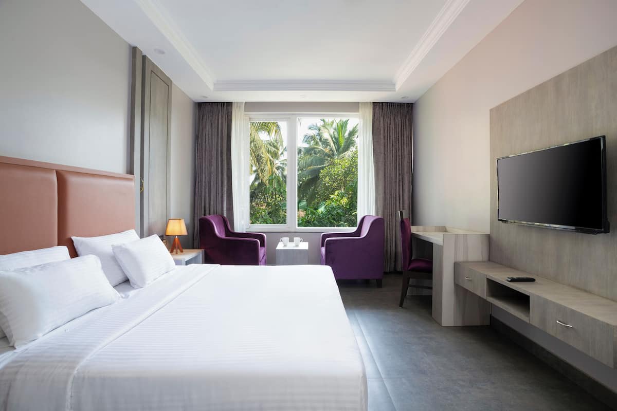 Quarto de hotel com uma cama grande, uma escrivaninha, uma TV e uma janela com vista para vegetação.